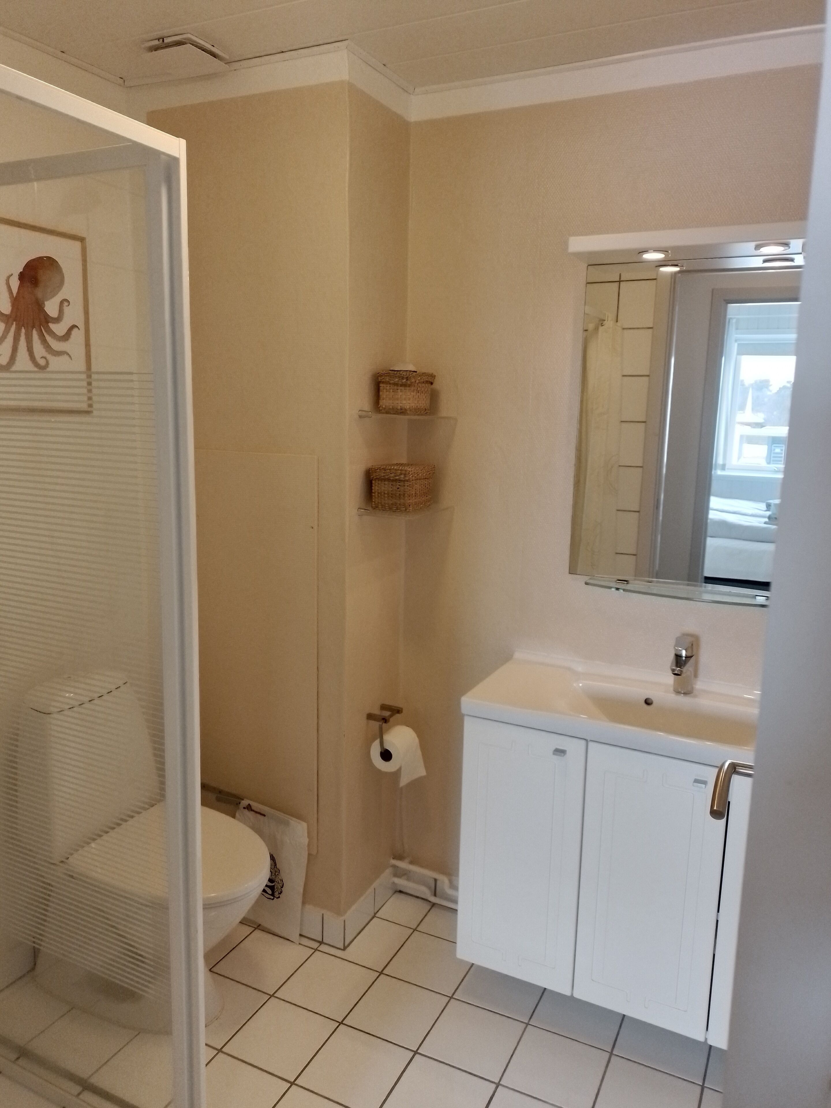 Casa de banho com sanita, lavatório e chuveiro. Existem duas cestas na parede e um rolo de papel higiénico.