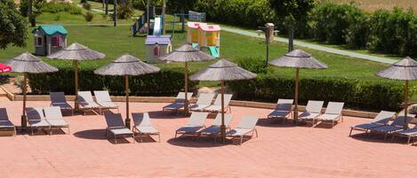 Área da piscina com espreguiçadeiras e guarda-sóis, cercada por vegetação e uma vista deslumbrante da água.