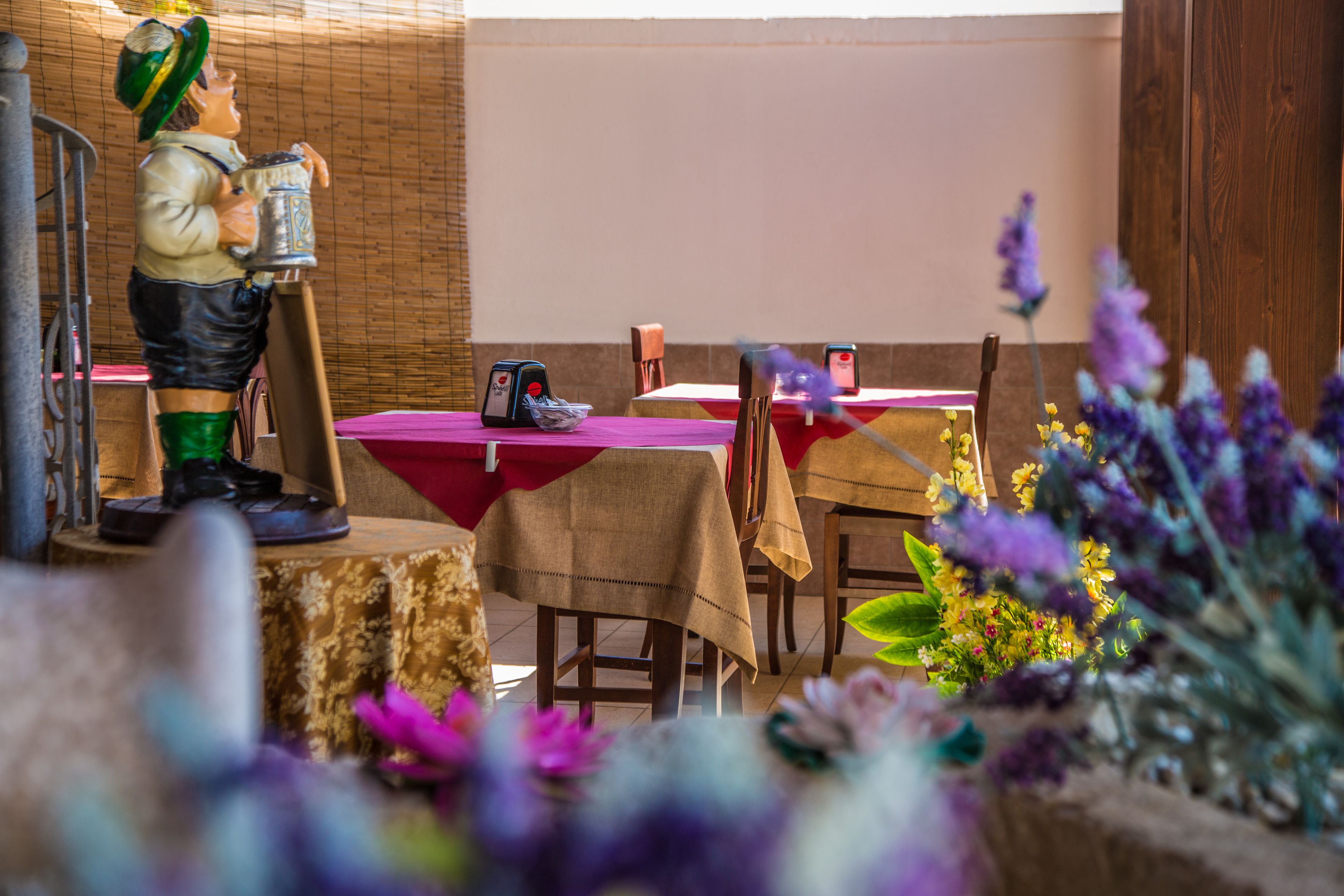 Restaurante com mesas cobertas por toalhas vermelhas, uma estátua de um homem segurando uma caneca de cerveja e diversos flores em primeiro plano.
