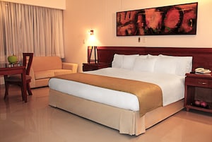 Quarto de hotel com uma cama grande, uma escrivaninha com abajur, uma cadeira e um quadro na parede.