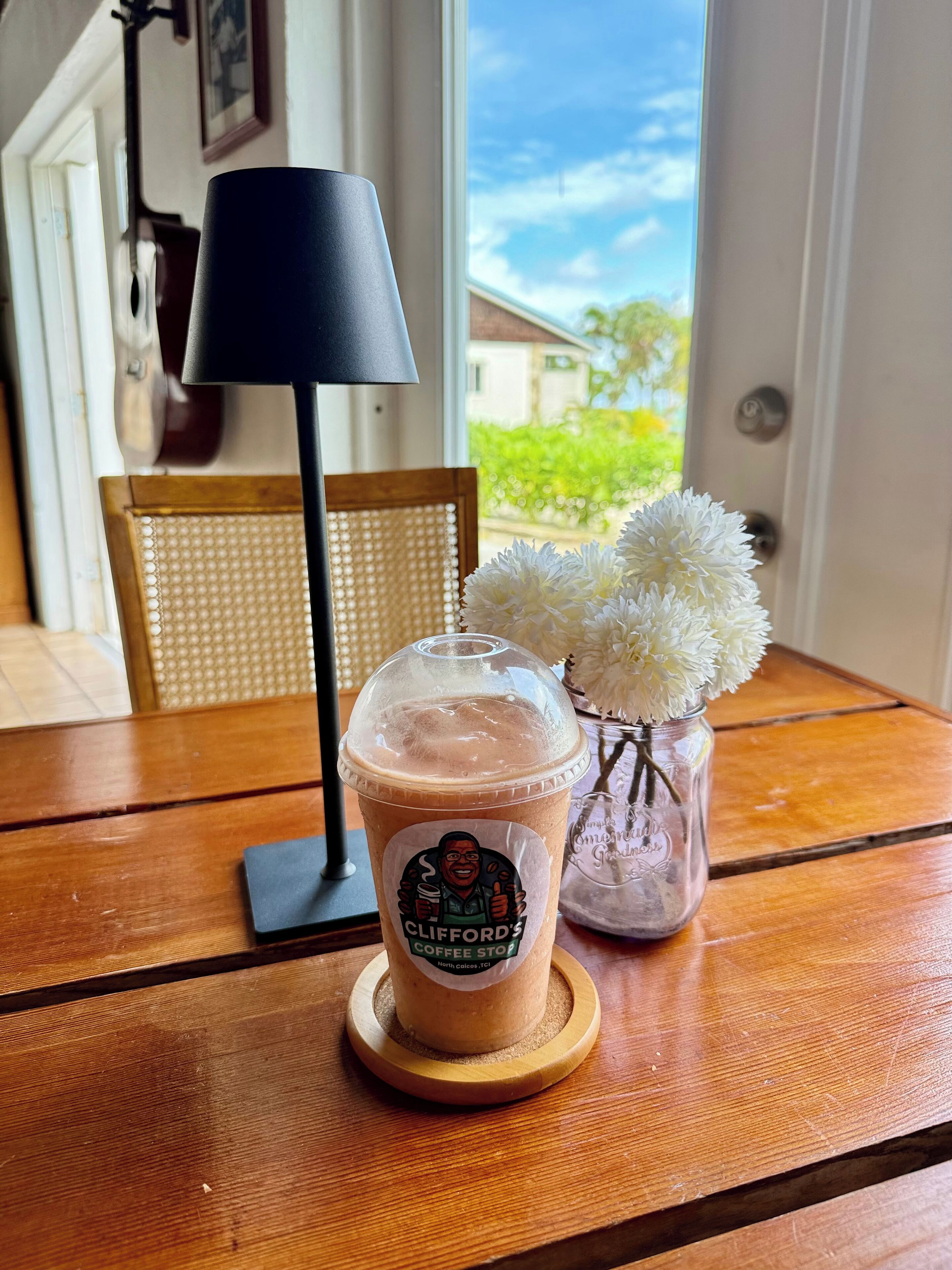 Uma xícara de café com tampa sobre um apoio, uma luminária e um vaso com flores em uma mesa de madeira.