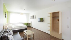 Quarto moderno e bem iluminado com cama, sofá, televisão e guarda-roupa de madeira.
