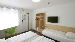 Quarto de hotel com duas camas, uma cadeira verde, uma estante de madeira, uma televisão e uma porta com aviso.