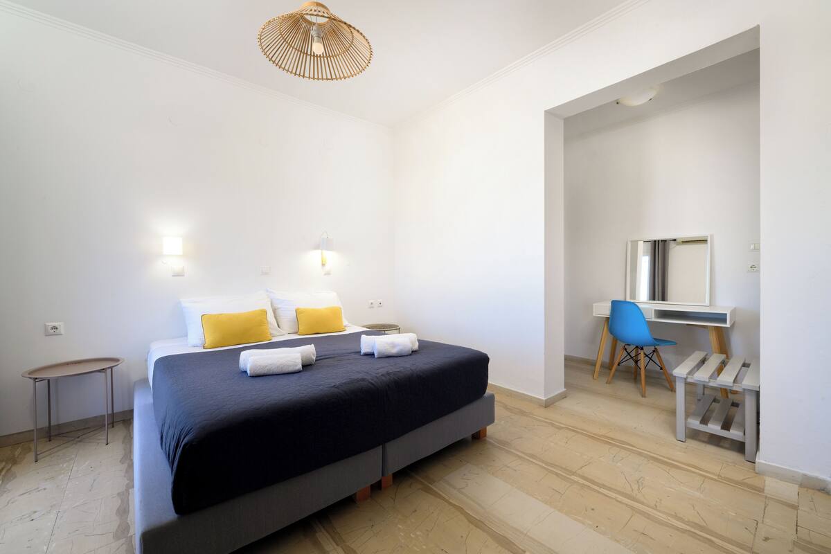 Quarto com cama, mesas de cabeceira, escrivaninha, cadeira e luminária suspensa.