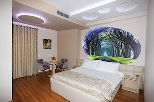 Quarto de hotel com cama, uma pequena mesa com cadeiras, mesa de cabeceira e um grande mural com tema de floresta na parede.