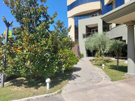 Um caminho leva a um edifício moderno com fachada de vidro e área de jardim ajardinada, com árvores e arbustos.