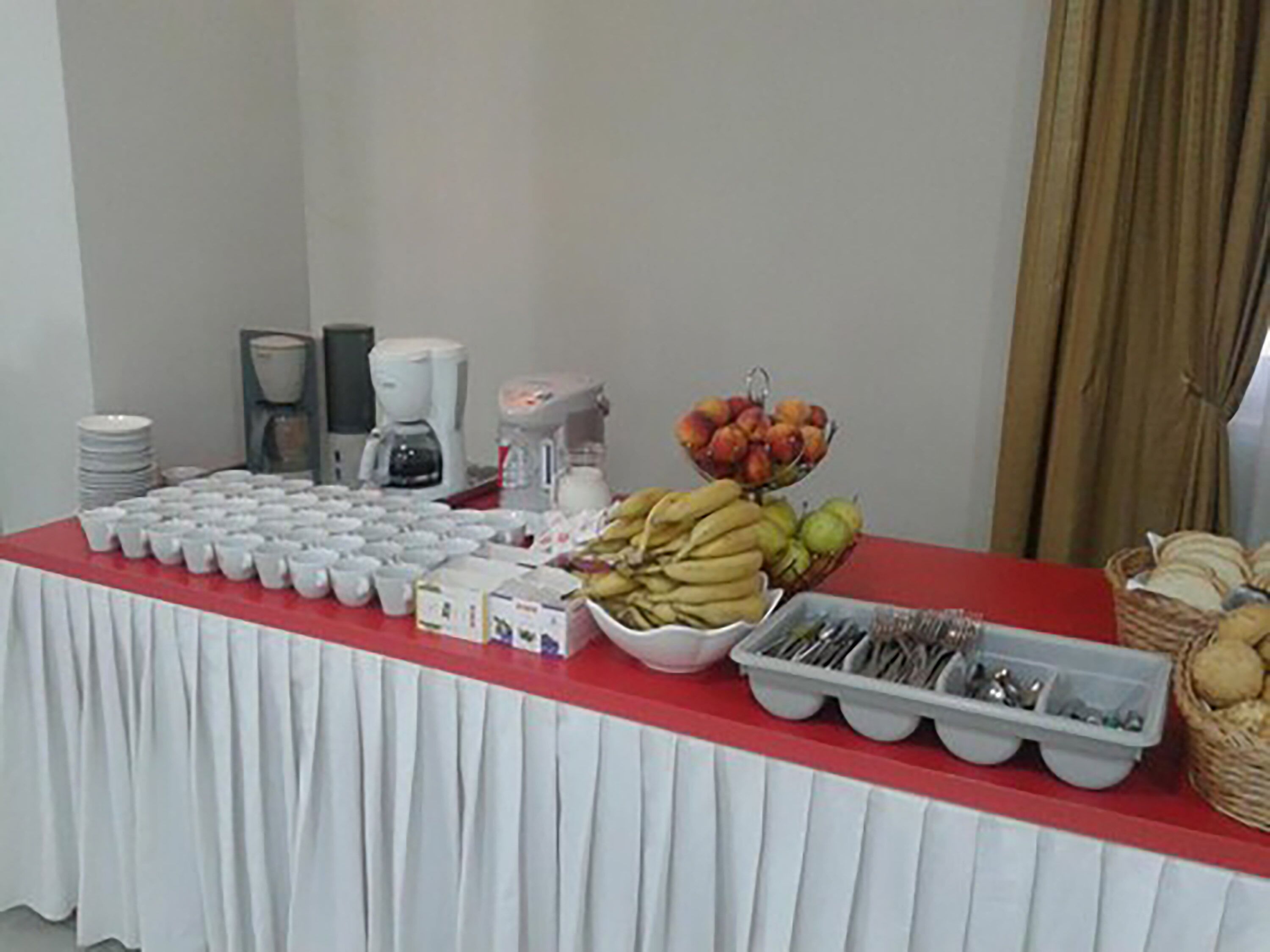 Uma mesa de buffet com diversos alimentos e utensílios, incluindo pratos, copos, bananas e uma saladeira.