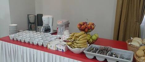 Uma mesa de buffet com diversos alimentos e utensílios, incluindo pratos, copos, bananas e uma saladeira.