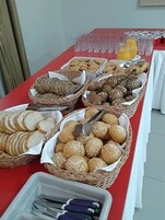 Uma mesa de buffet com diversos pães, pãezinhos e doces em cestas, além de um recipiente com utensílios e um jarro de suco de laranja.