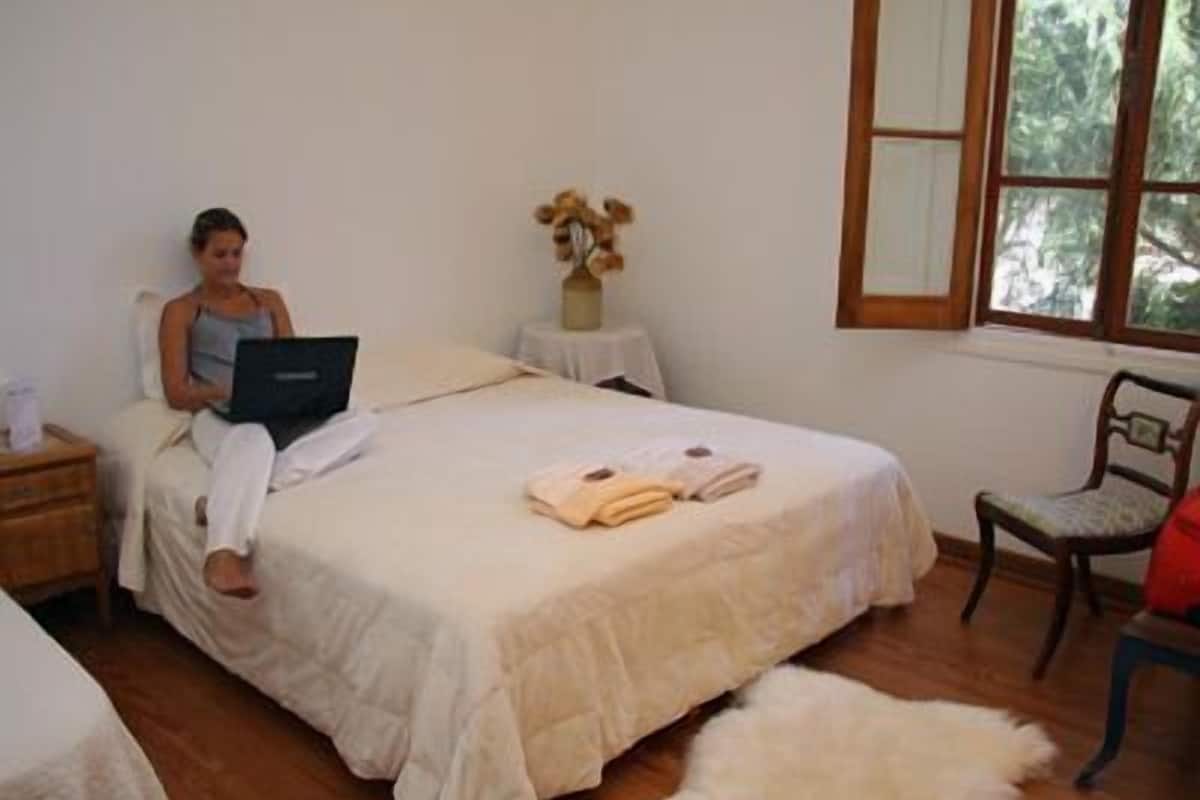 Quarto com cama, laptop, um vaso com flores, cadeira e janela.