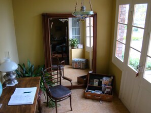 Quarto com escrivaninha de madeira, cadeira e uma pequena mesa com abajur. Há uma mala e uma caixa no chão. Uma porta dá acesso a outro quarto com cadeira e mesa.