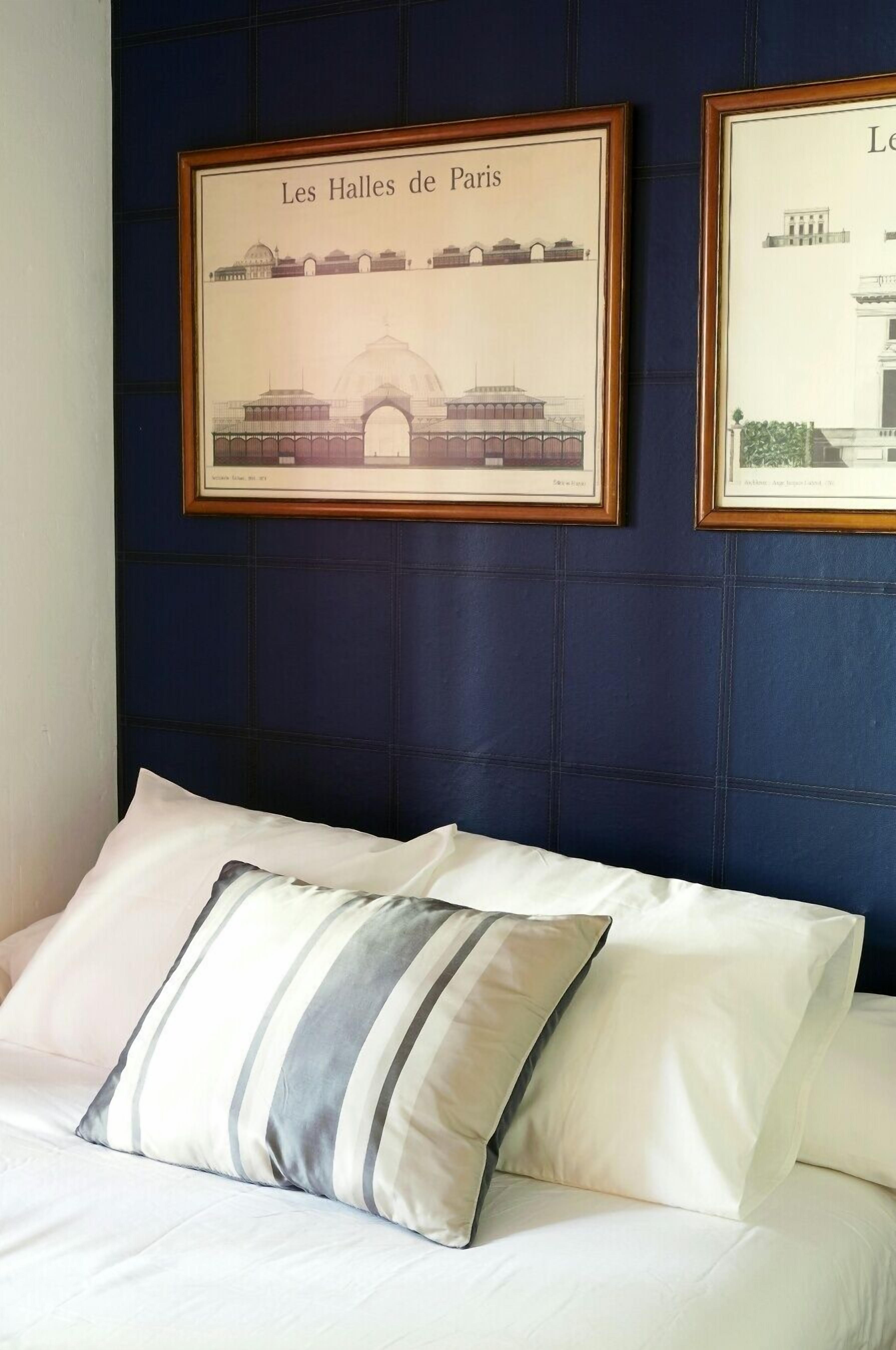 Uma cama com roupa de cama branca e almofadas listradas, uma parede azul escura com um quadro de pontos turísticos de Paris e outro quadro de um edifício.