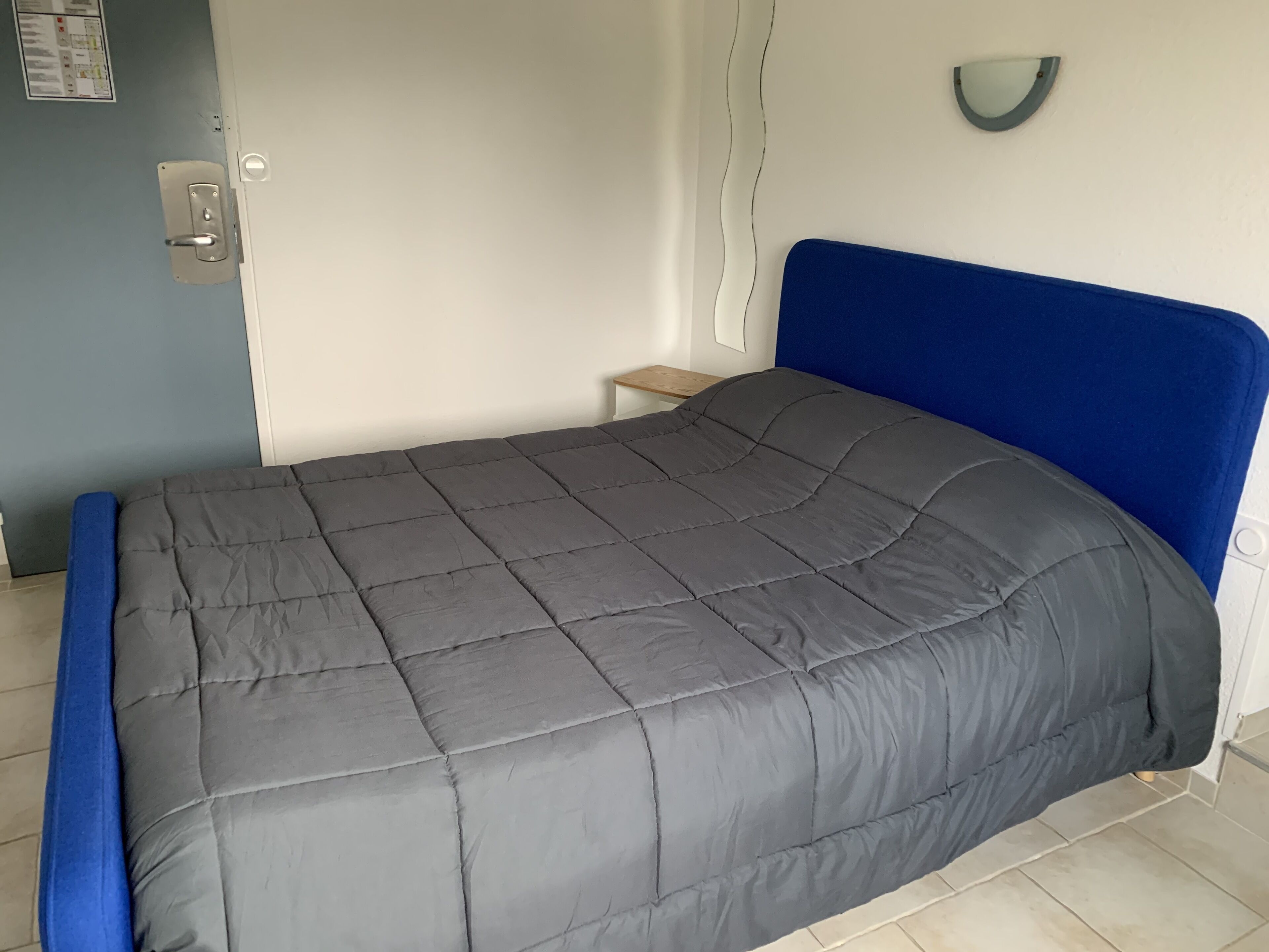 Quarto com uma cama de solteiro, uma mesa de cabeceira pequena e um espelho fixado na parede.