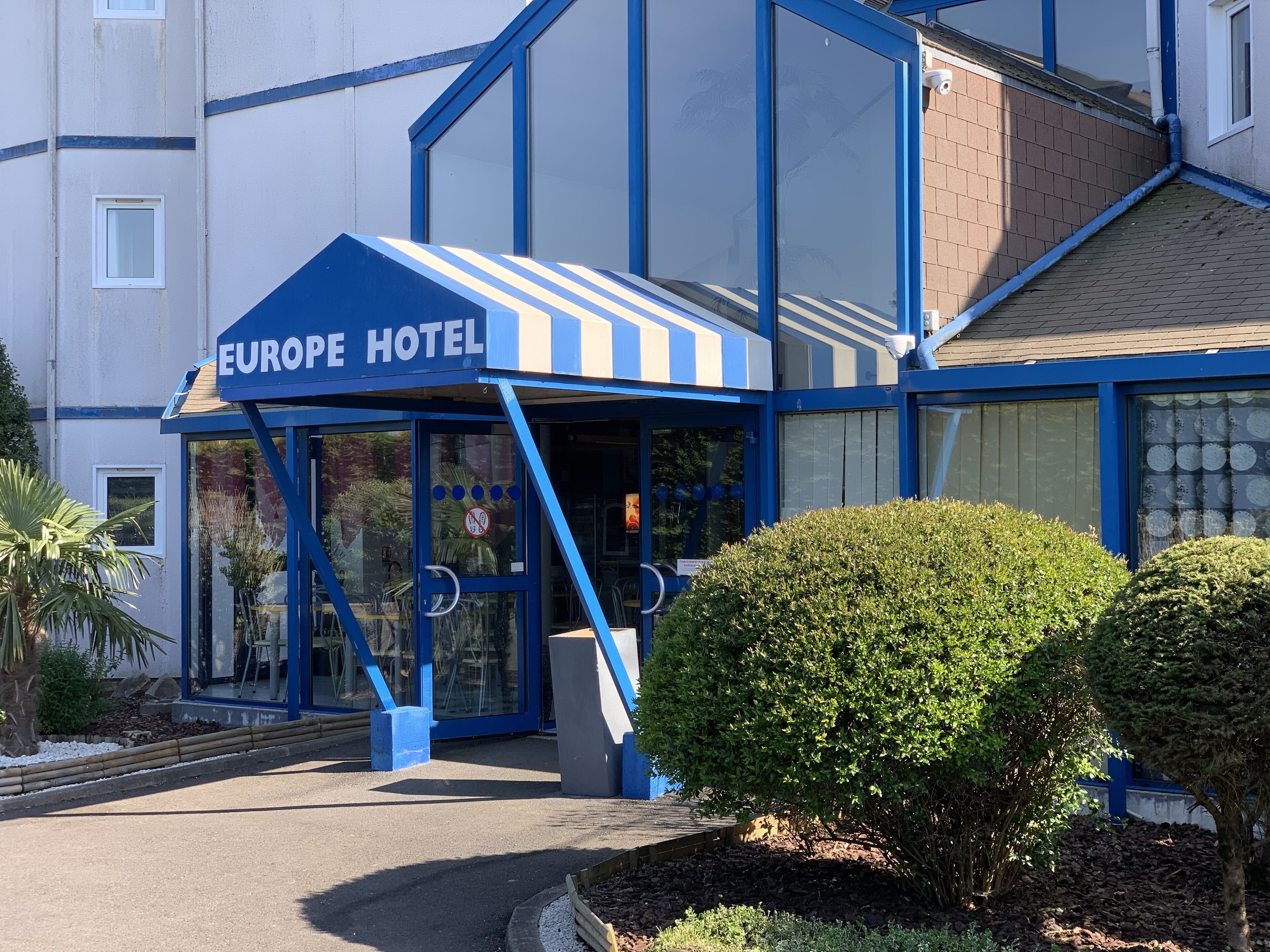 Exterior do Hotel Europa, com toldo azul e branco listrado, entrada de vidro e parede de tijolos adjacente ao edifício.