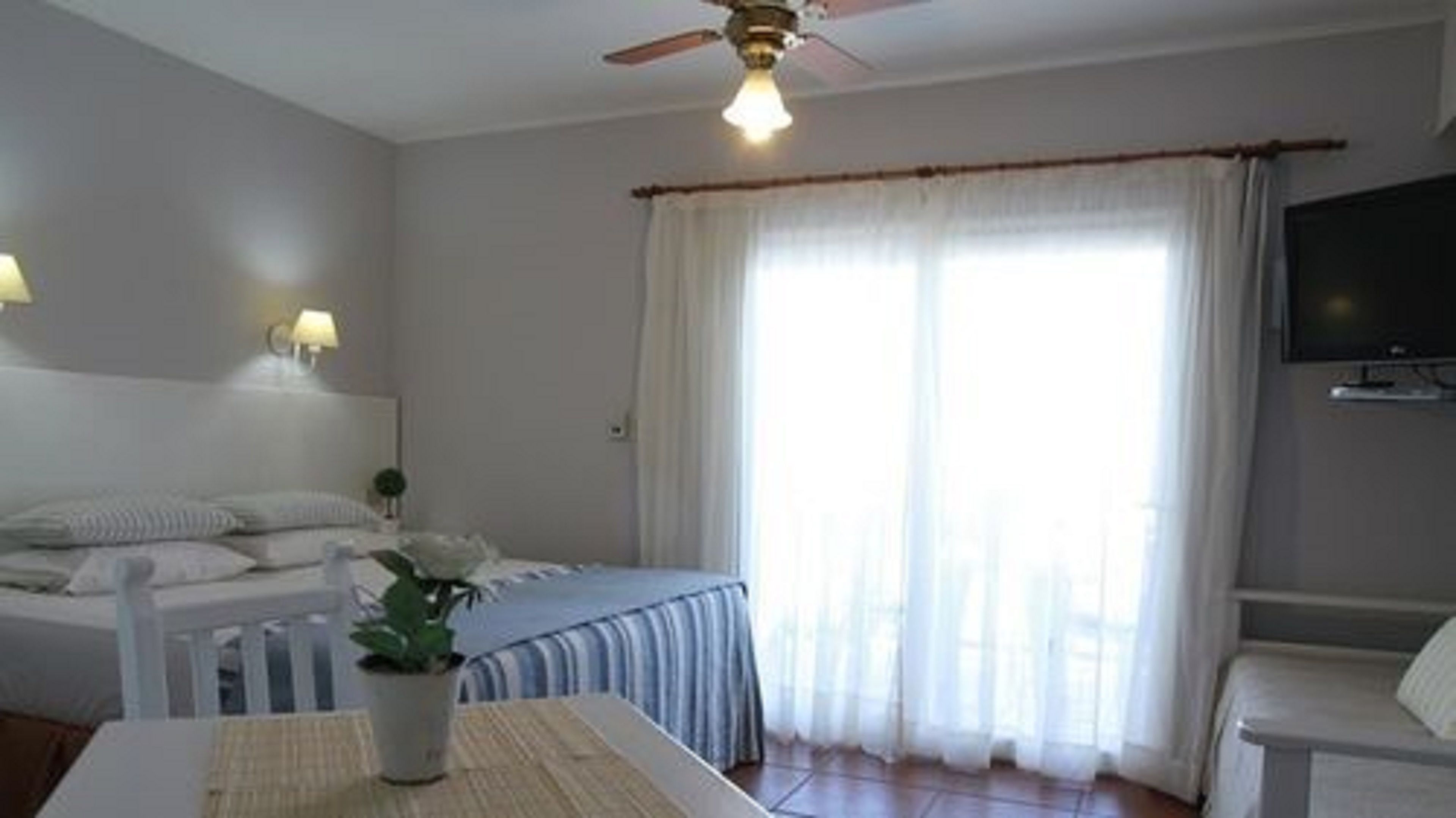Quarto de hotel com uma cama, uma escrivaninha, uma cadeira, uma televisão e um ventilador de teto.