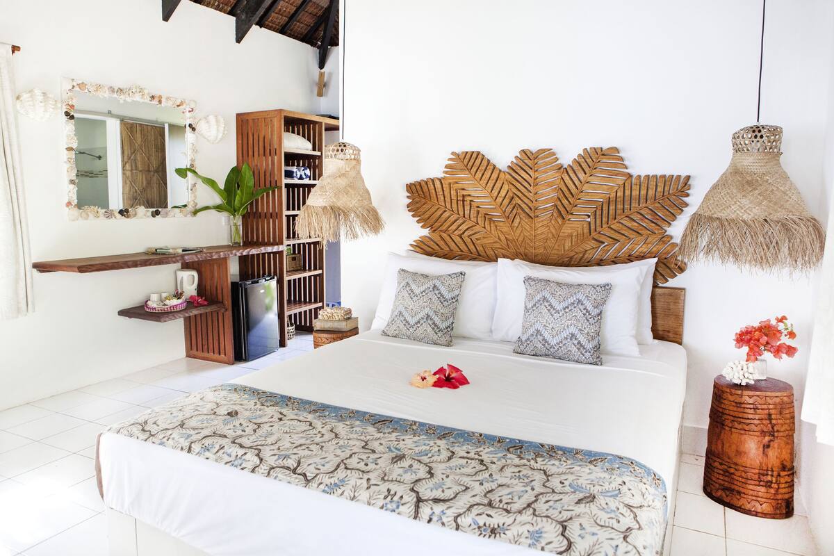 Quarto com uma cama grande, cabeceira de madeira, espelho, prateleira com objetos decorativos e luminária pendente.