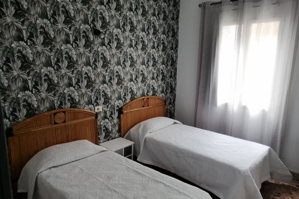 Quarto com duas camas de solteiro, cabeceira de madeira, mesa de cabeceira pequena e janela com cortinas.