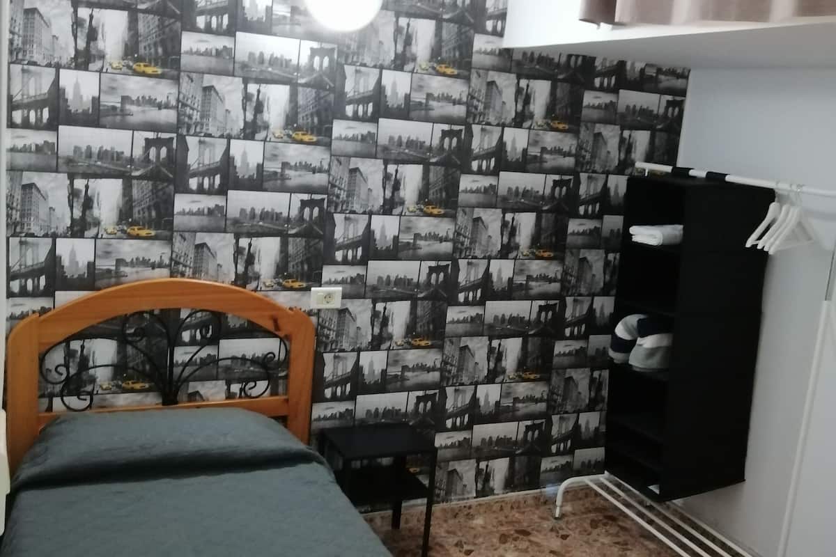 Quarto com cama, uma mesinha, um guarda-roupa e parede com padrão de paisagem urbana.