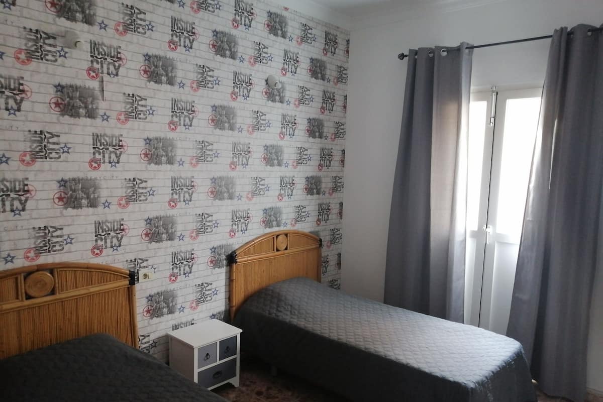 Quarto com duas camas de solteiro, uma pequena mesa de cabeceira, parede com papel de parede estampado e uma janela com cortinas.