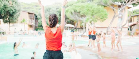 Uma pessoa de camisa vermelha e shorts pretos ao lado de uma piscina, com os braços levantados, rodeada por outras pessoas e árvores.