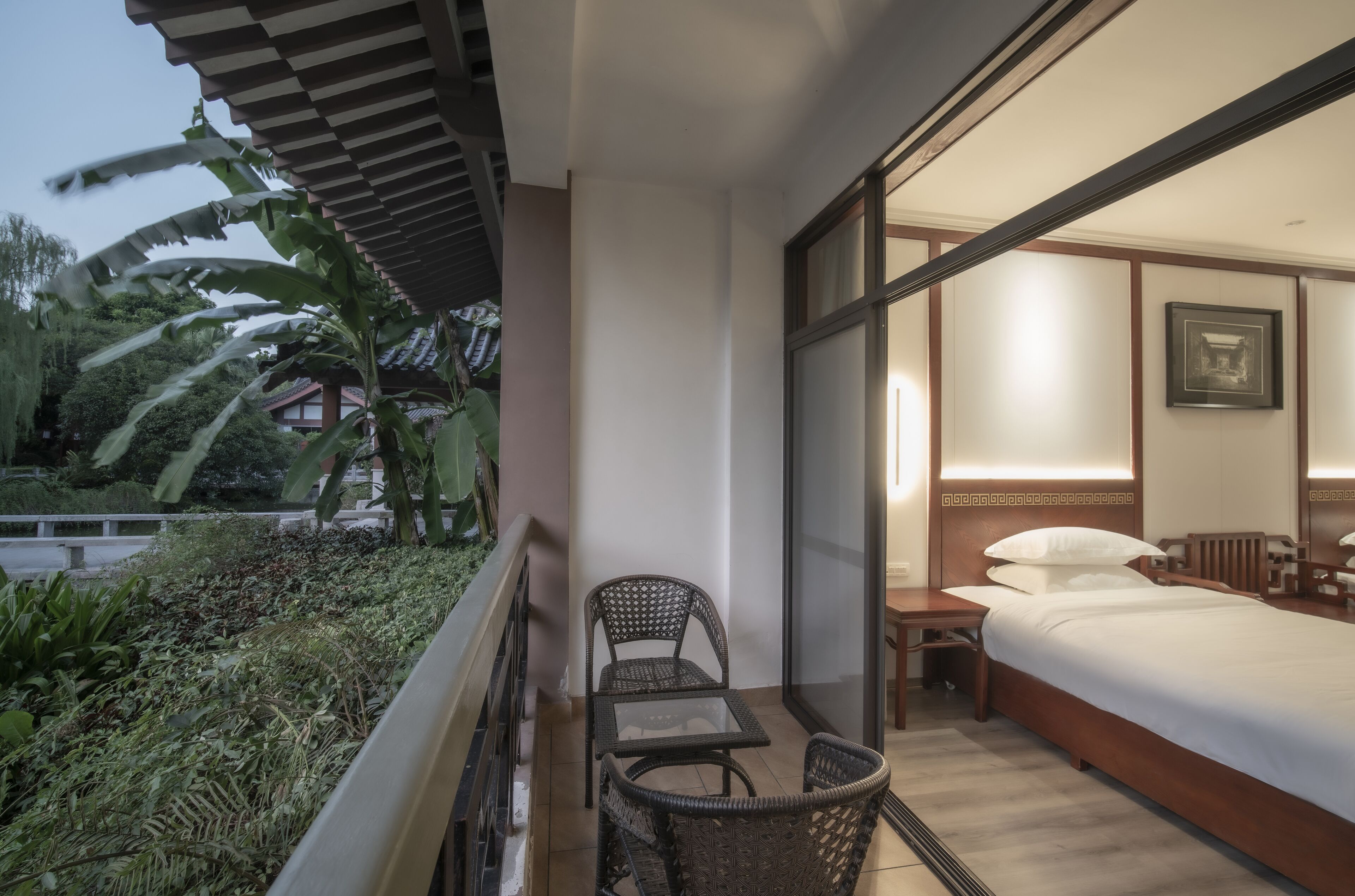 Quarto de hotel com uma cama, uma mesinha, uma cadeira e uma varanda com plantas.