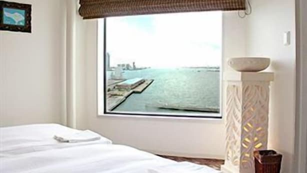 Quarto de hotel com uma cama, janela com vista para a água, um objeto decorativo na parede e uma mesinha com uma tigela.