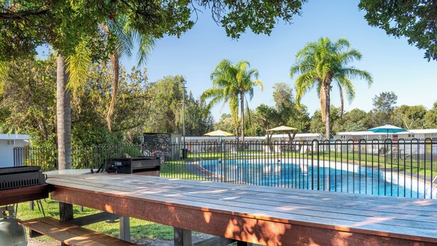 Um deck de madeira com piscina, cercado por palmeiras e um muro.