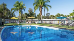 Piscina com espreguiçadeiras e guarda-sóis, cercada por palmeiras e uma área murada com edifícios ao fundo.
