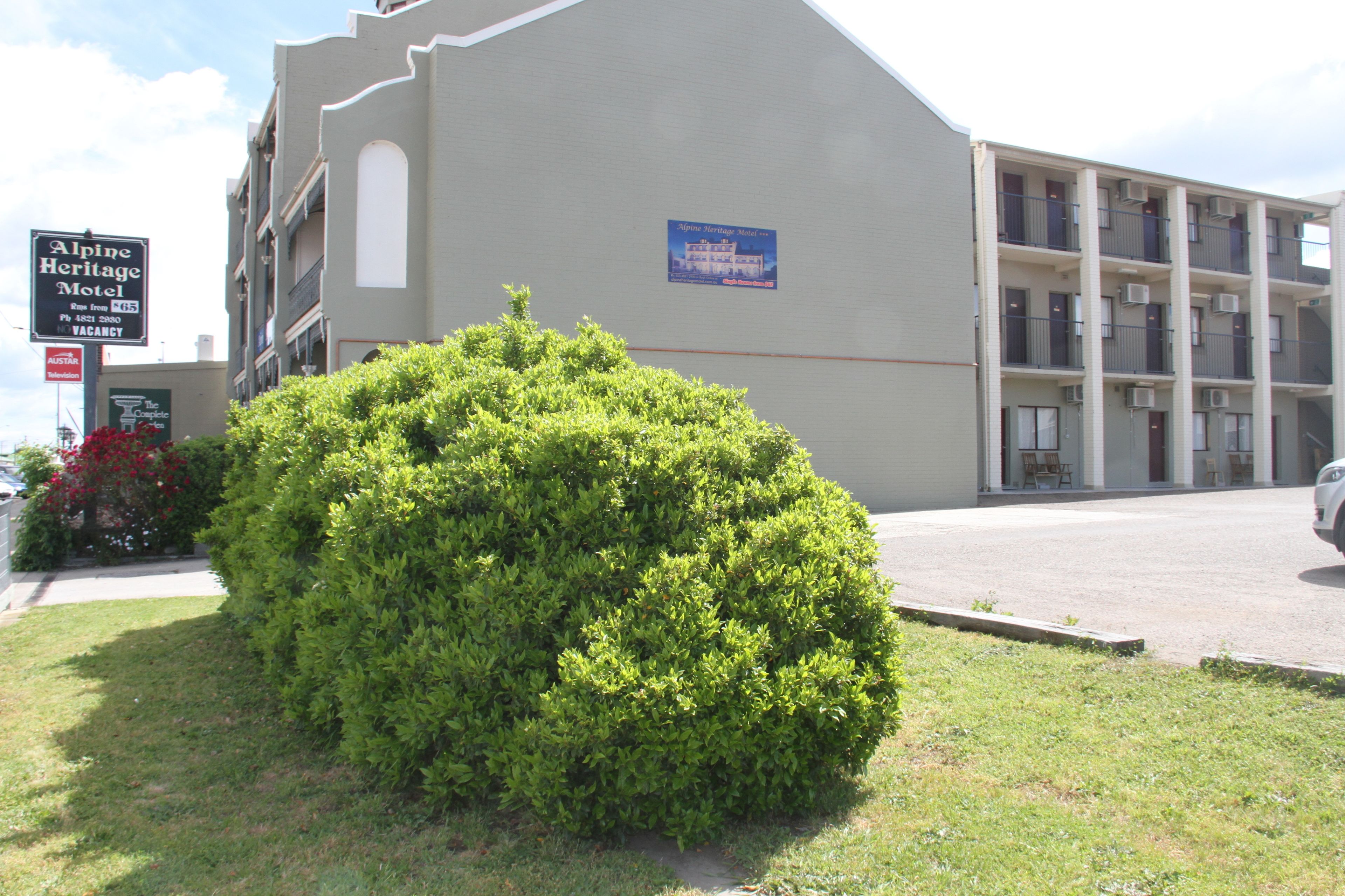 Um edifício com a placa do Alpine Heritage Motel, um arbusto grande na frente e uma estrutura de vários andares com varandas.