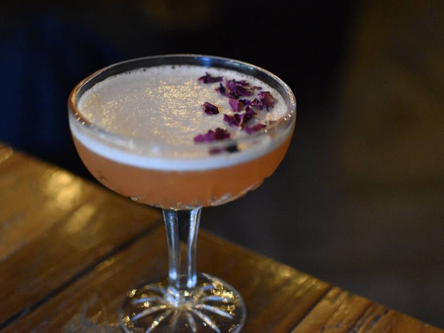 Um cocktail num copo com espuma por cima e guarnição roxa sobre uma superfície de madeira.