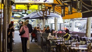 Uma área de refeições ao ar livre com mesas e cadeiras, um letreiro de pizzaria e uma van estacionada.