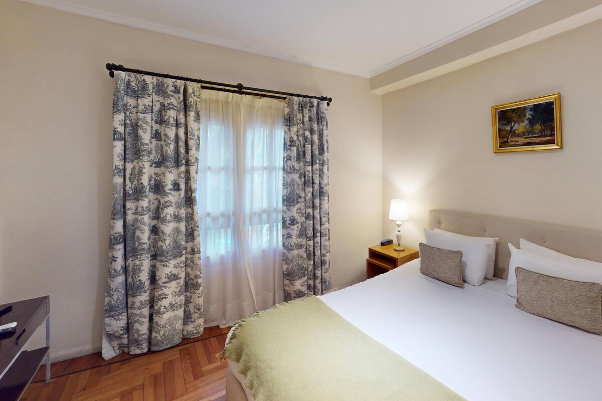 Quarto de hotel com uma cama, mesa de cabeceira com abajur, pintura na parede e cortina com padrão floral.