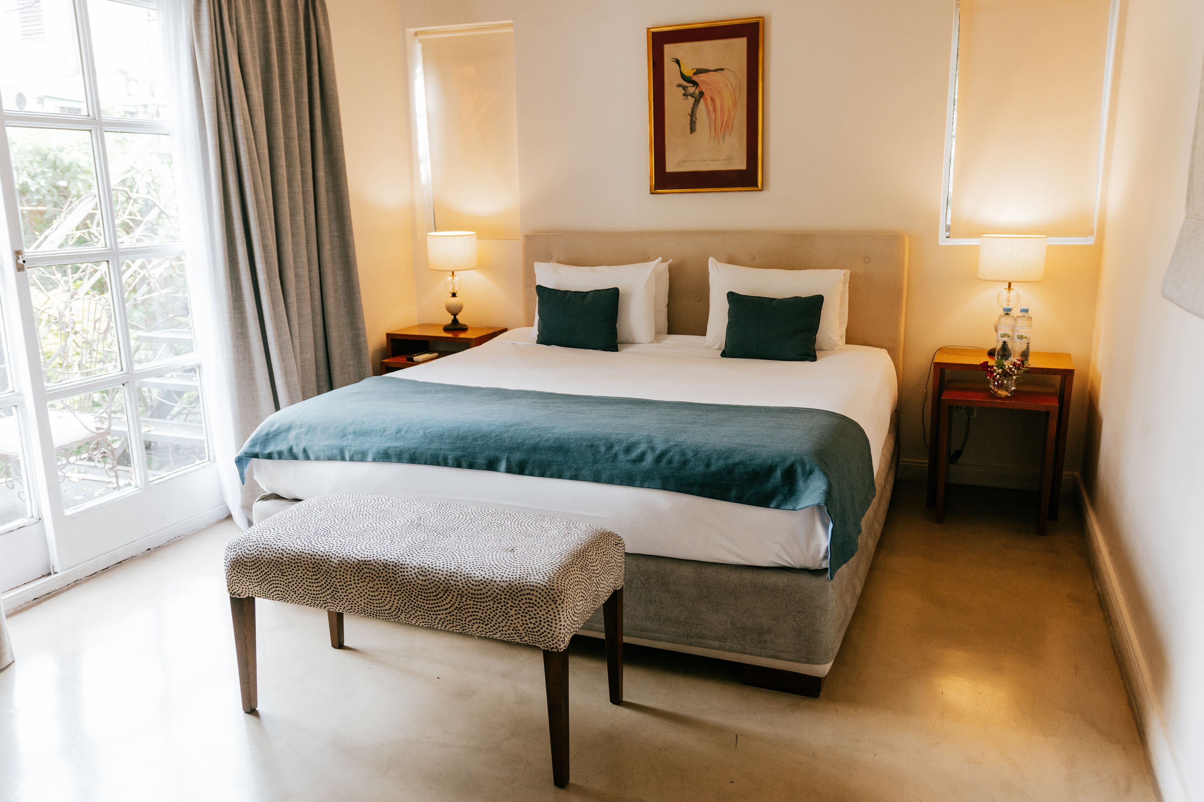 Cama bem arrumada com colcha azul e cinza, duas almofadas e um banquinho combinando na frente. Há duas mesas de cabeceira com luminárias, uma obra de arte emoldurada na parede e uma janela grande com cortinas.
