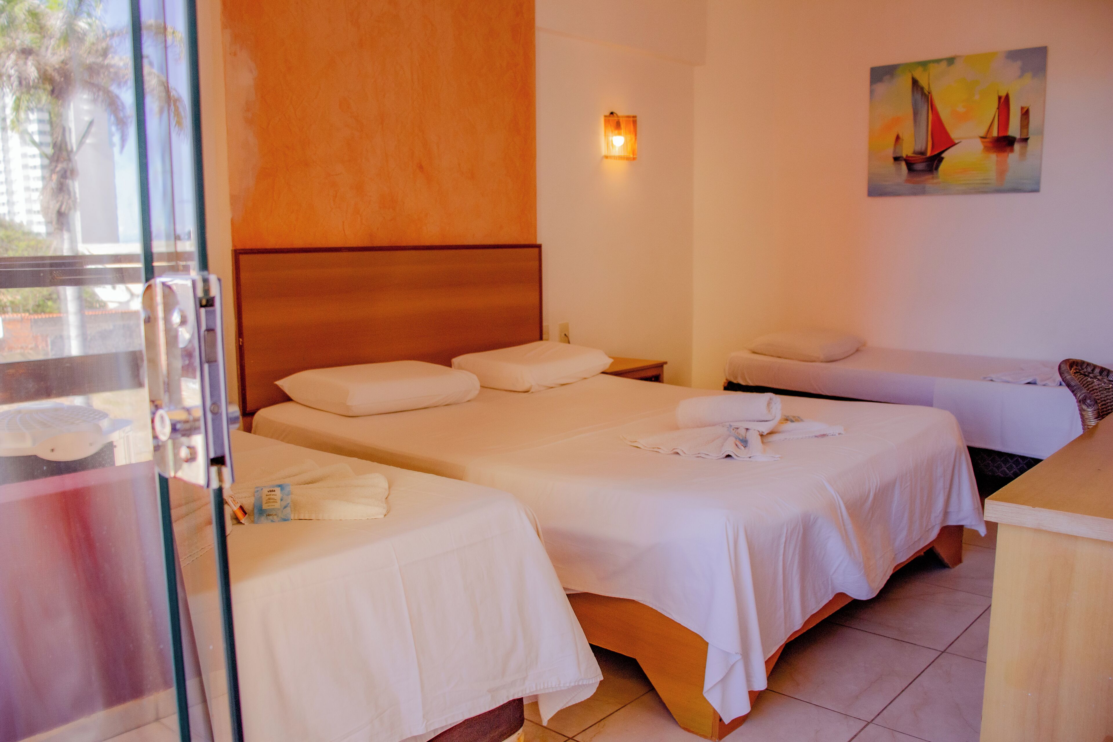 Quarto de hotel com duas camas, uma cabeceira de madeira, um quadro na parede e uma janela com vista para palmeiras.