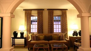 Uma sala de estar aconchegante com um sofá, duas poltronas e uma mesa de centro. Há duas luminárias em mesas laterais, e o espaço apresenta um design arquitetônico clássico com teto abobadado e colunas.