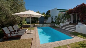 Piscina retangular com borda de tijolos, cercada por uma área gramada com quatro espreguiçadeiras de madeira e um guarda-sol.