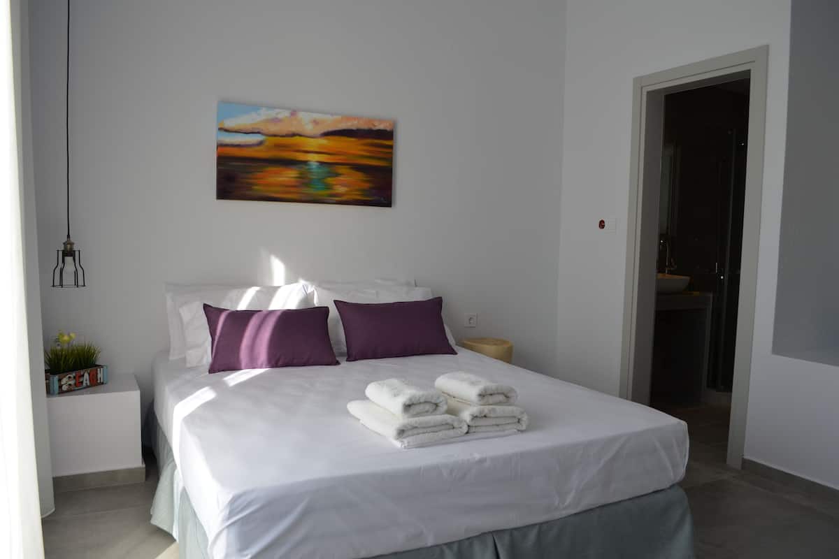 Cama bem arrumada com lençóis brancos e almofadas roxas, um quadro na parede e uma pequena planta na mesa de cabeceira.