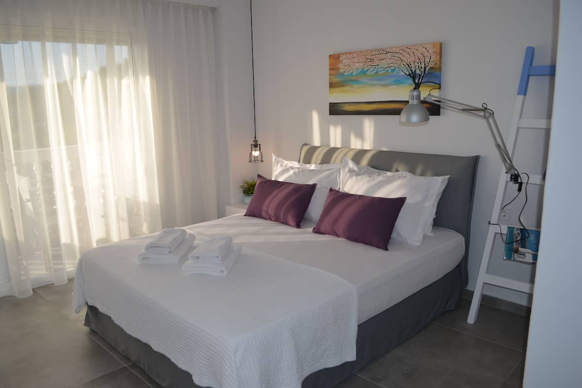 Cama bem arrumada com almofadas brancas e roxas, abajur e pintura na parede.