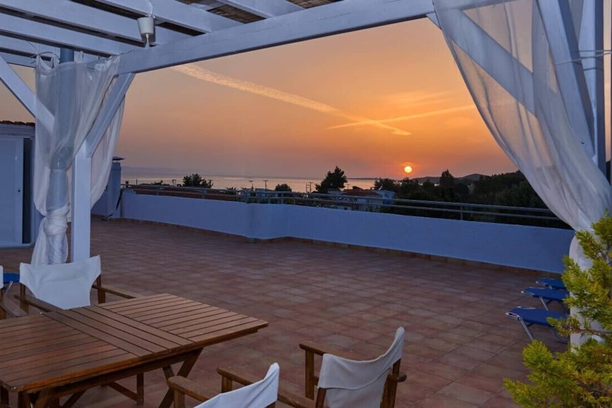 Terraço com mesa e cadeiras de madeira, vista do pôr do sol sobre o mar e uma estrutura de pérgula branca.