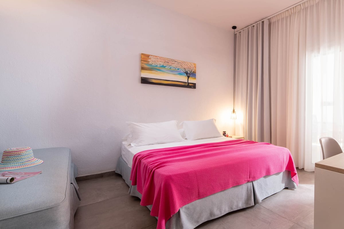 Quarto de hotel com cama, secretária, cadeira, um quadro na parede e janela com cortinas.
