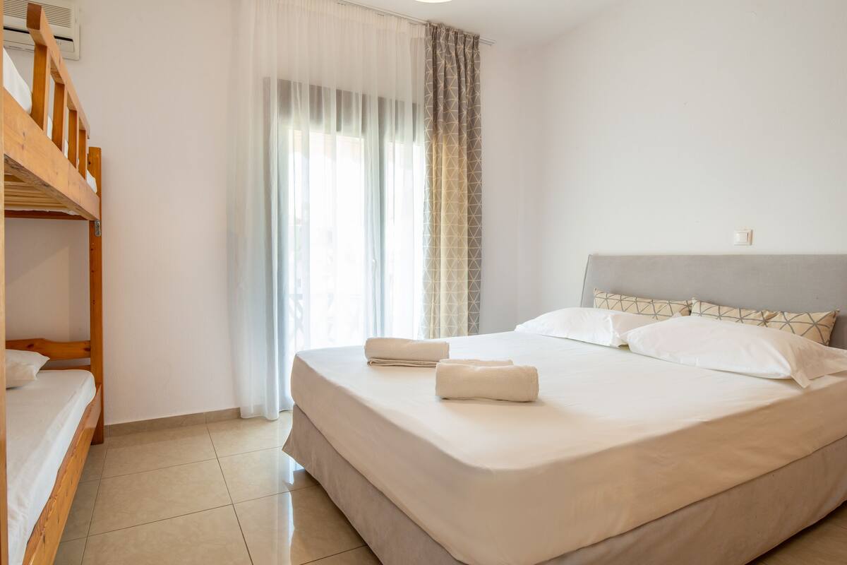 Quarto de hotel com uma cama grande, uma beliche de madeira, cortinas leves e chão de azulejos.