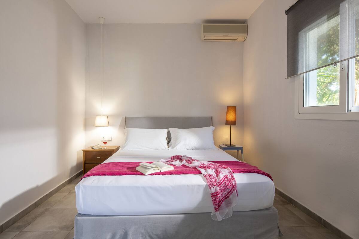 Cama bem arrumada com lençóis brancos e um cobertor estampado em rosa e branco. Há uma mesa de cabeceira com uma luminária e um criado-mudo com um telefone.