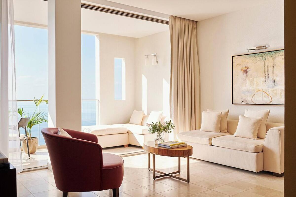 Uma sala de estar moderna com uma poltrona vermelha, um sofá bege e uma mesa de centro. Há uma grande janela que oferece luz natural e vista para o exterior.