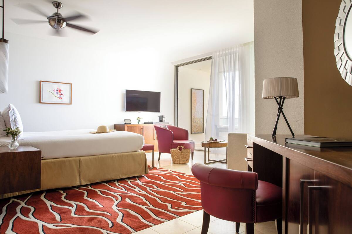 Quarto de hotel com cama, escrivaninha, cadeira, televisão e ventoinha no teto.