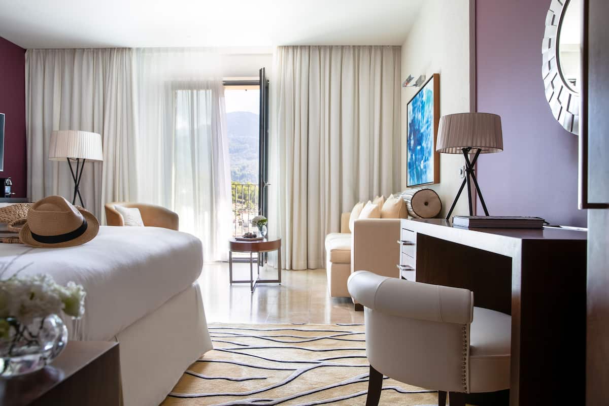Quarto de hotel com uma cama, uma escrivaninha, uma cadeira, um abajur, um relógio e vista para o exterior.
