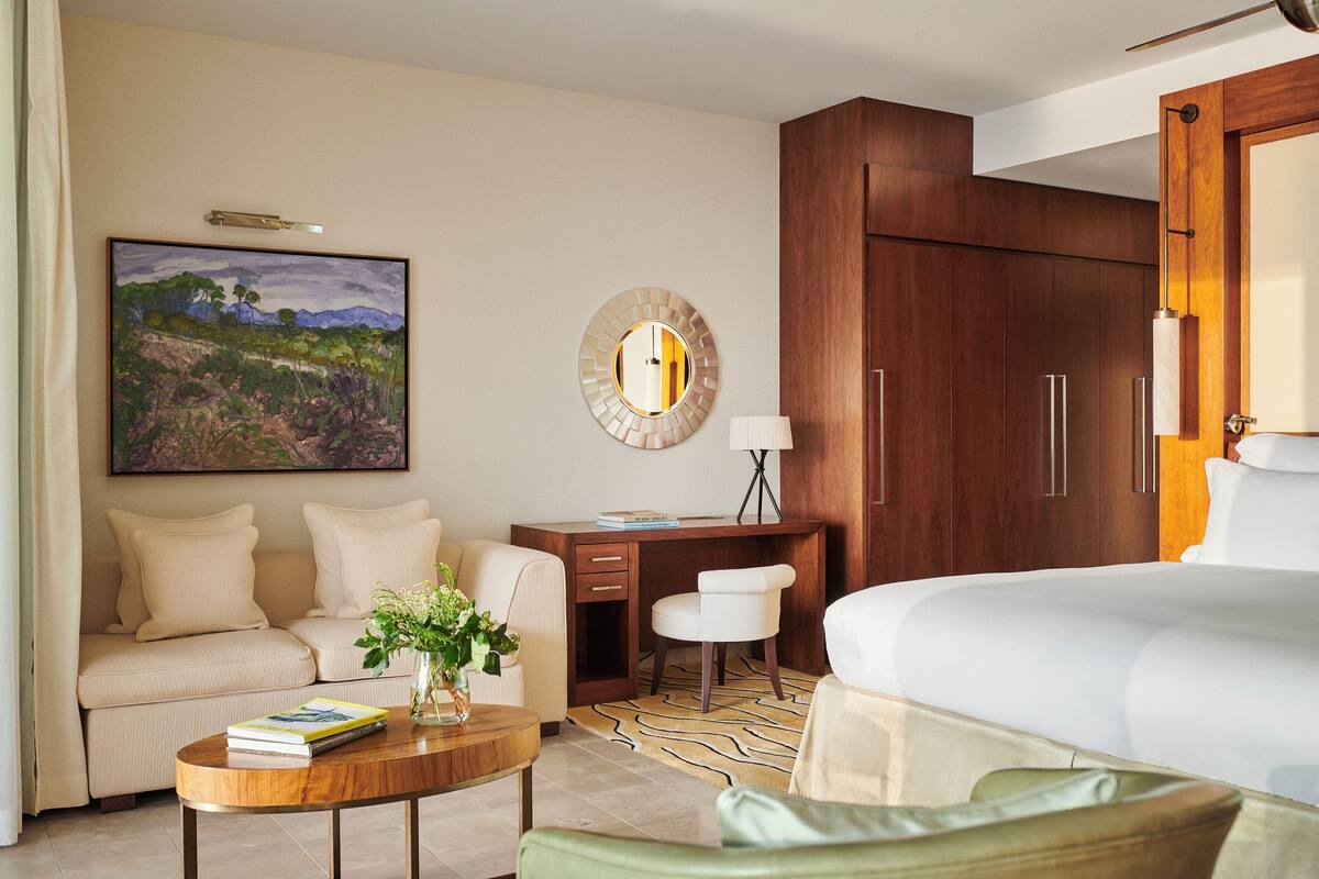 Quarto de hotel com uma cama, uma escrivaninha, uma cadeira, um sofá, um espelho redondo e um quadro na parede.