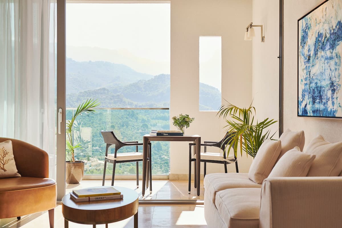 Uma sala de estar moderna com um sofá, uma mesa de centro e duas cadeiras. É visível uma paisagem montanhosa através de uma grande janela.