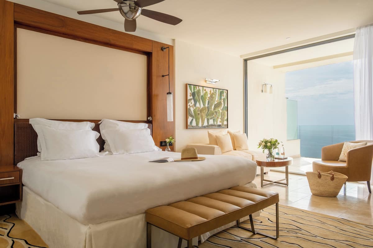 Quarto de hotel com uma cama grande, um ventilador de teto, uma pintura, uma varanda com vista e uma área de estar com mesa e cadeiras.