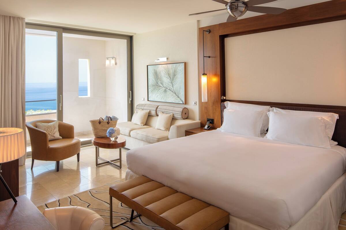 Quarto de hotel com uma cama grande, um sofá, uma poltrona, uma mesinha e vista para o oceano.