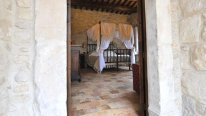 Um quarto com paredes de pedra, uma cama sob teto de madeira, piso de pedra e um arco de pedra.
